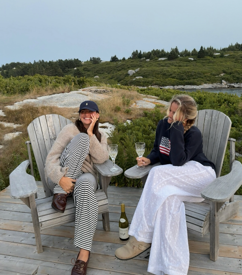 Two women relaxing outdoors in casual cozy outfits%2C one wearing a Ralph Lauren navy cap%2C beige fuzzy sweater%2C striped pants%2C and brown loafers%2C the other in a navy sweatshirt with an American flag%2C white wide-leg pants%2C and beige boots%2C seated on wooden Adirondack chairs with wine glasses and bottle nearby.